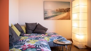 Um quarto com uma cama, almofadas, uma mesa redonda com um livro, um abajur fixado na parede e uma pintura de uma praia.
