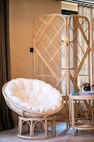 Uma cadeira de vime com assento e encosto acolchoados, uma pequena mesa com um livro e um objeto decorativo, e um divisório de bambu em um quarto com piso de azulejos e cortina.