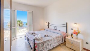 Quarto com uma cama, uma mesa de cabeceira com um frasco e flores, e vista para a praia por uma porta deslizante.