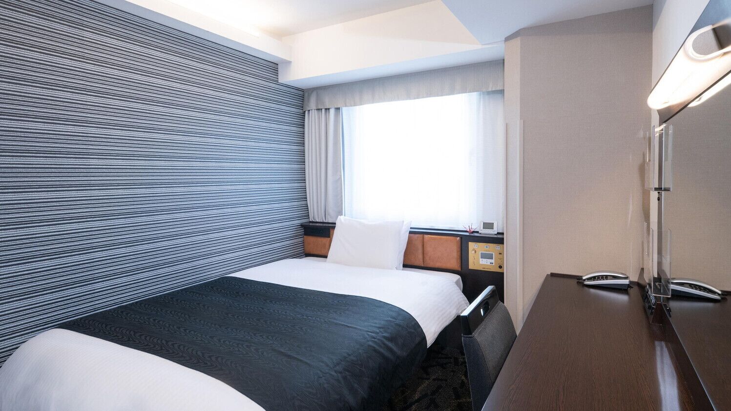 Quarto de hotel com cama, escrivaninha, cadeira, janela com cortinas e parede com padrão listrado.