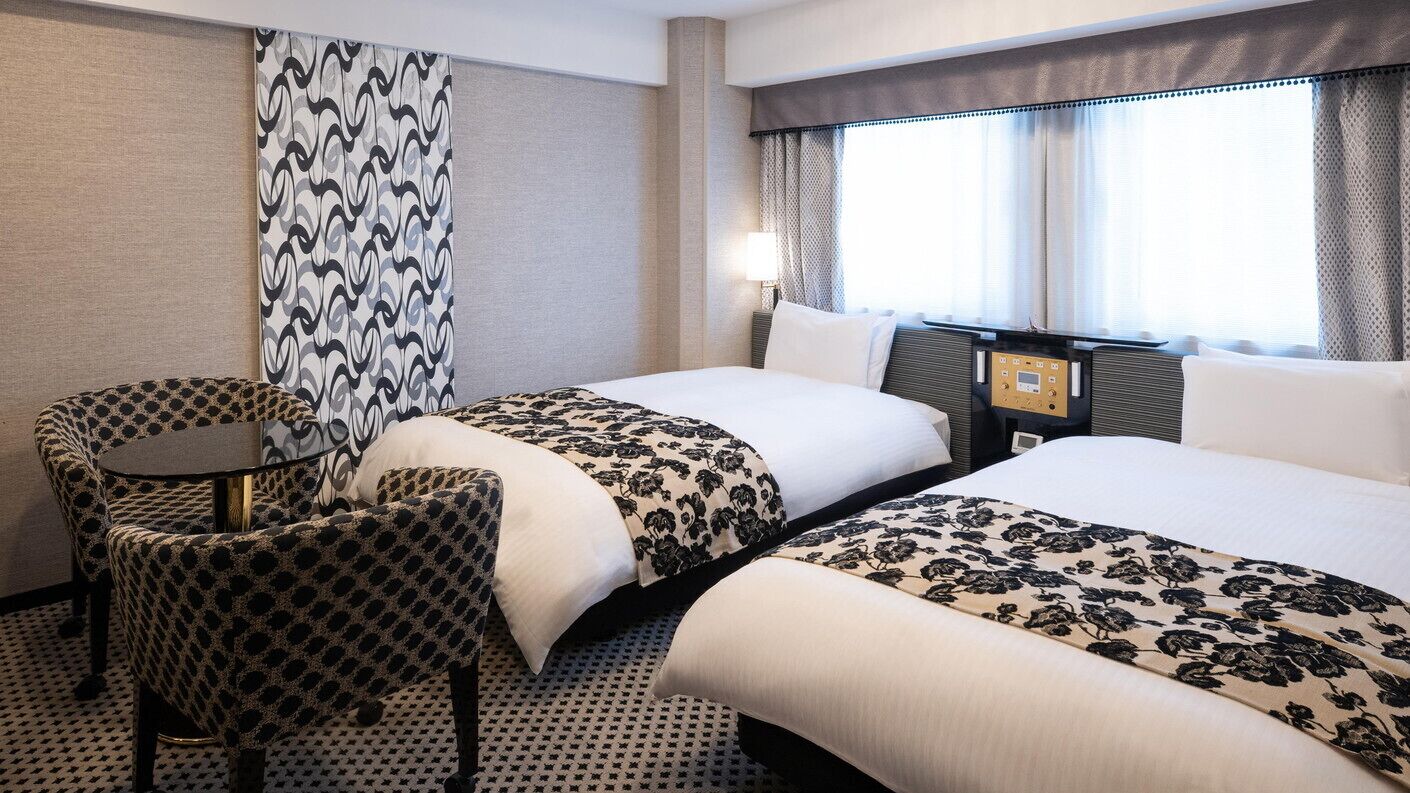 Quarto de hotel com duas camas, uma cadeira, uma mesa de cabeceira e uma janela com cortinas.