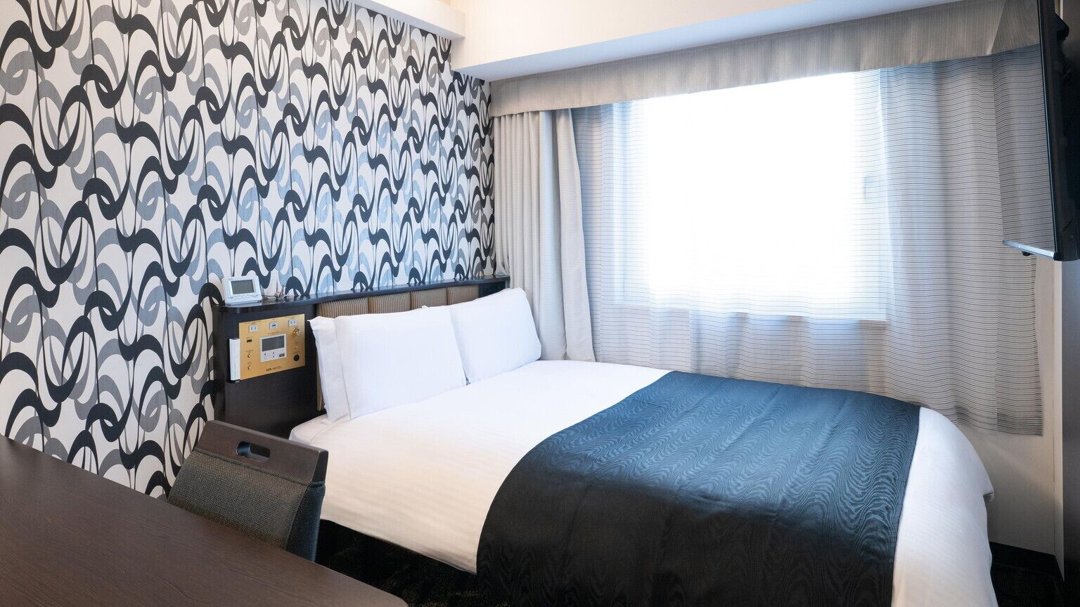 Quarto de hotel com cama, mesa de cabeceira, painel de controle e janela com cortinas.