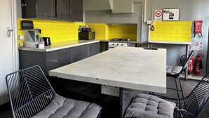 Uma cozinha moderna com uma ilha central, painel de azulejos amarelos e armários pretos. Há duas cadeiras pretas com almofadas cinzas, um micro-ondas e um fogão a gás.