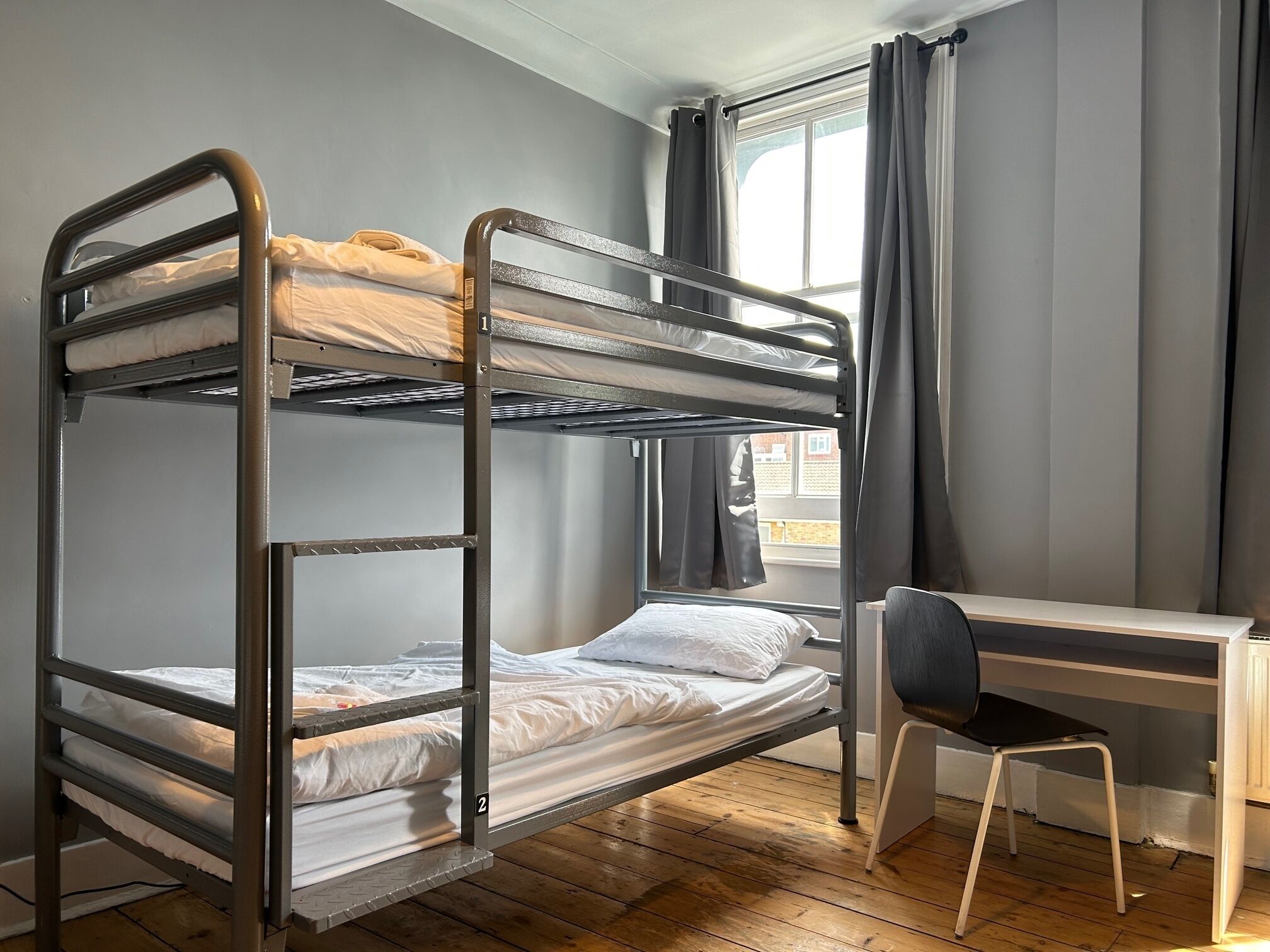 Quarto com beliche, escrivaninha e cadeira, janela com cortinas e vista para o exterior.