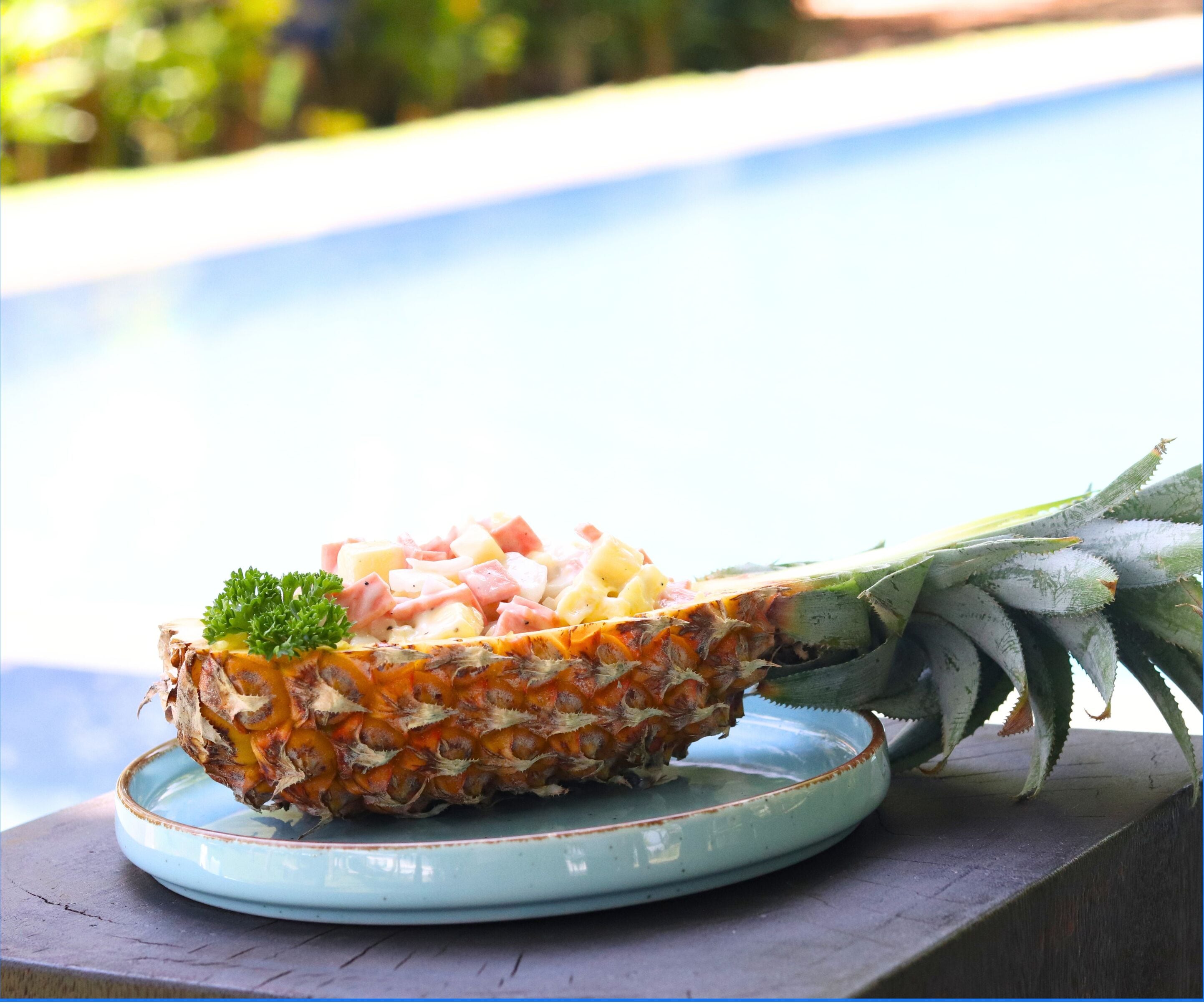 Uma abacaxi com uma tigela de salada de frutas picada sobre um prato azul, próximo a uma piscina.