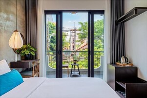 Quarto de hotel com uma cama grande, uma escrivaninha com luminária, uma mesinha com uma planta e uma varanda com vista para áreas verdes e edifícios.