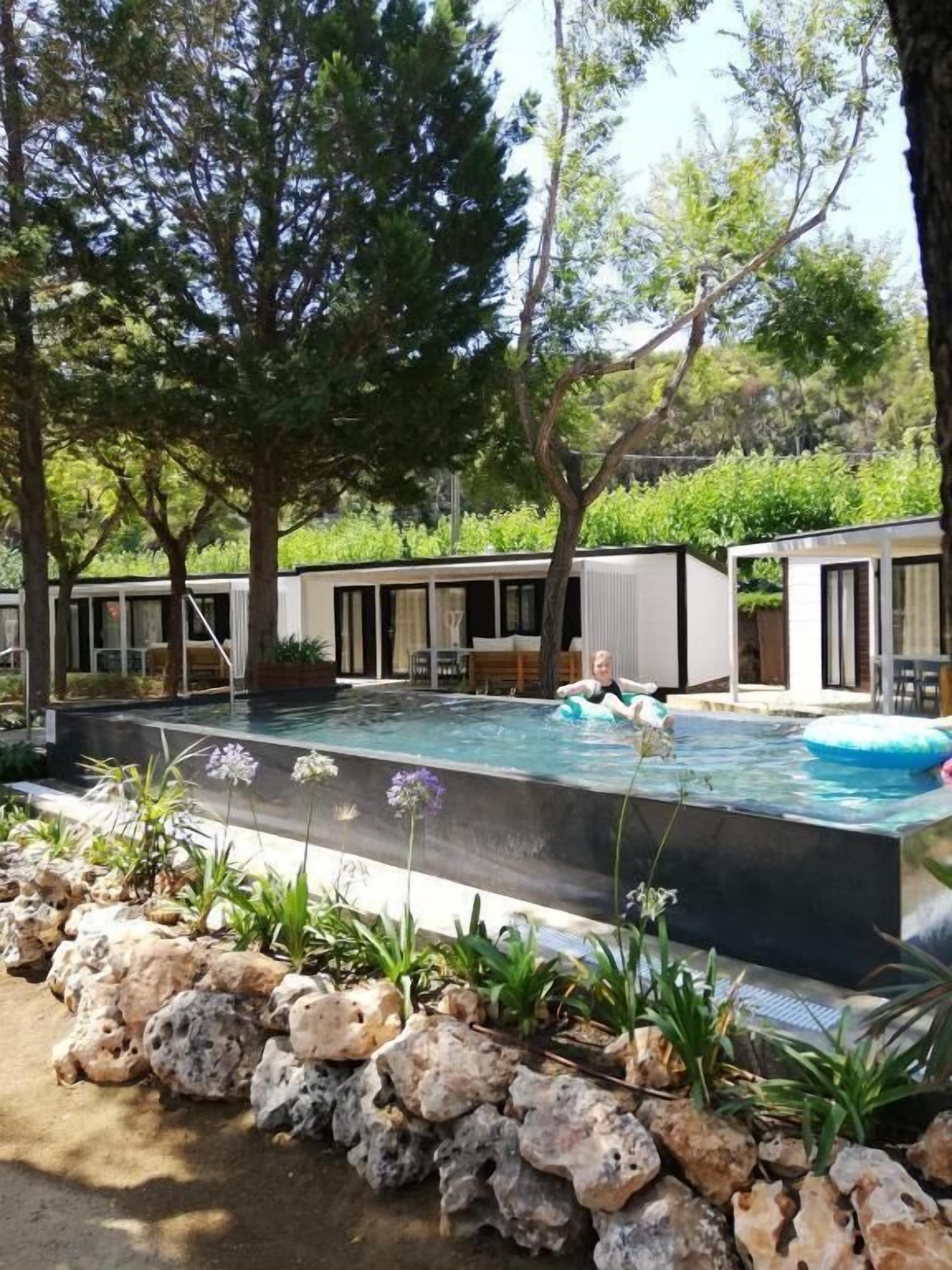 Piscina exterior moderna com borda de pedra, rodeada por vegetação exuberante e árvores.