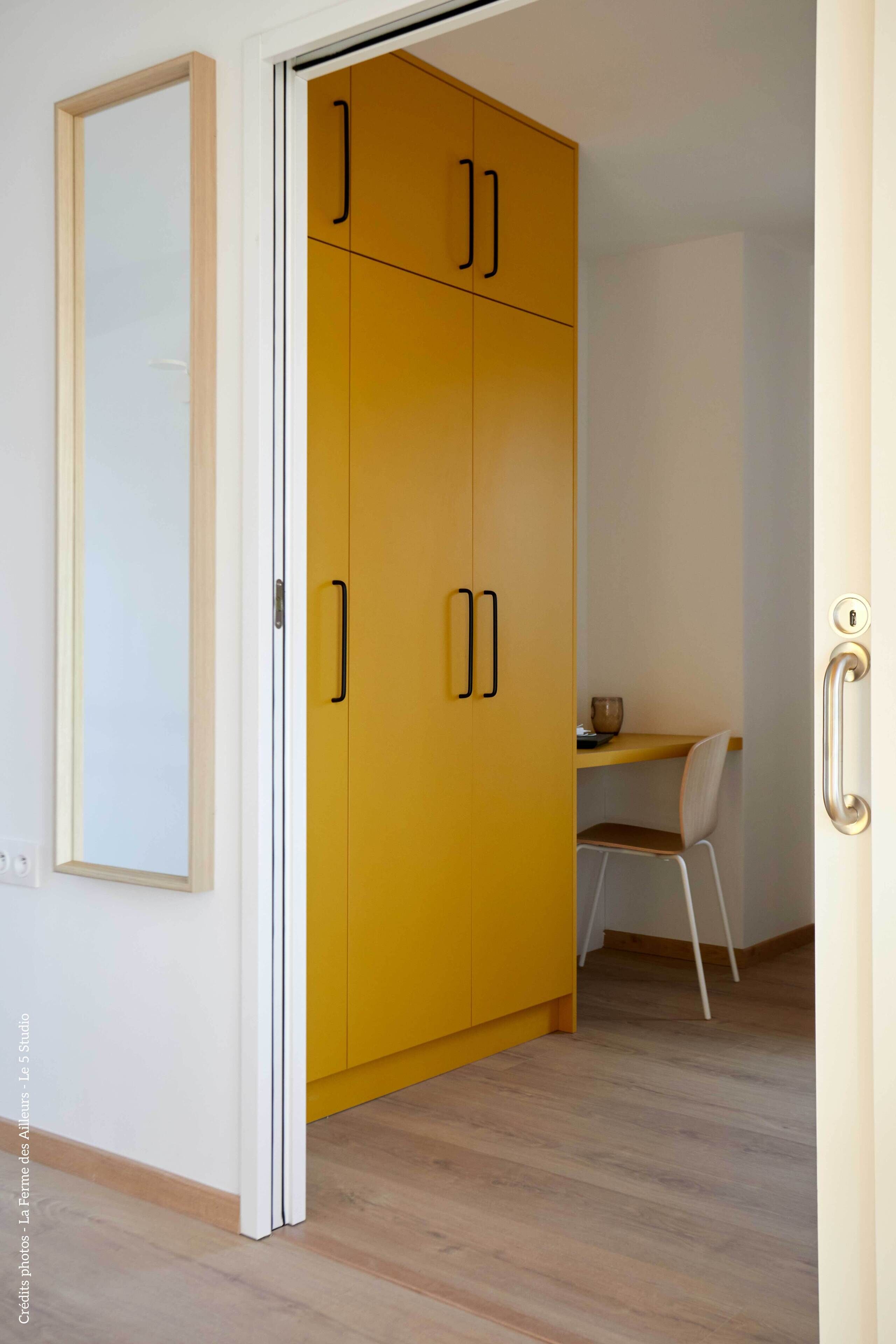 Quarto com piso de madeira, guarda-roupa amarelo com puxadores pretos, uma cadeira branca e uma mesinha com uma tigela.