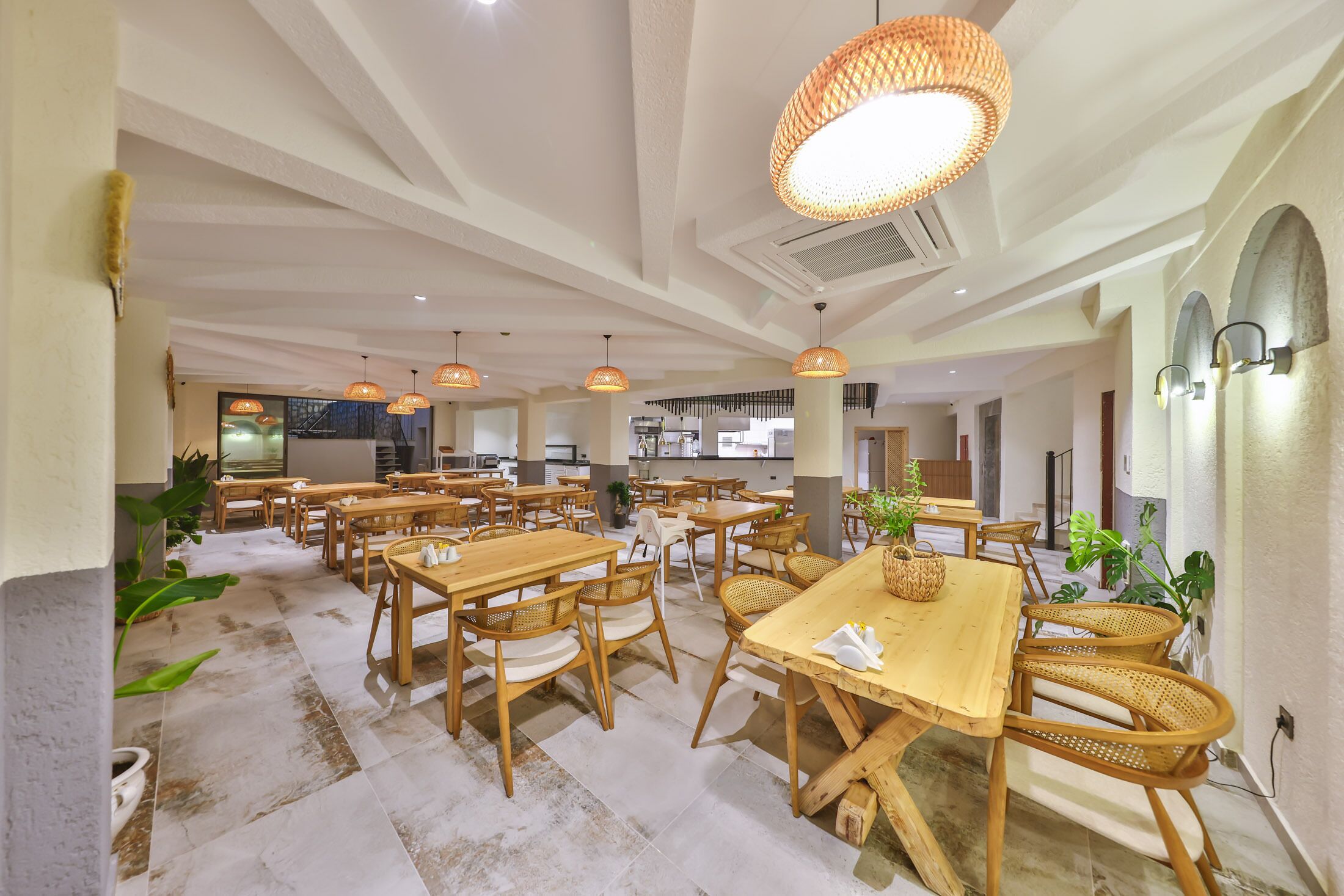 Uma sala de jantar ampla com várias mesas e cadeiras de madeira, luminárias pendentes de vime no teto e plantas em vasos.