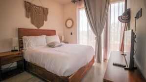 Quarto com cama de madeira, roupa de cama branca, mesa de cabeceira com abajur, televisão sobre suporte de madeira e janela com cortinas.