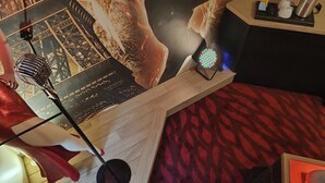 Quarto com piso de madeira, tapete vermelho estampado, parede decorativa com um pôster, pequena mesa com um chaleira e um objeto decorativo que se assemelha a um pássaro.