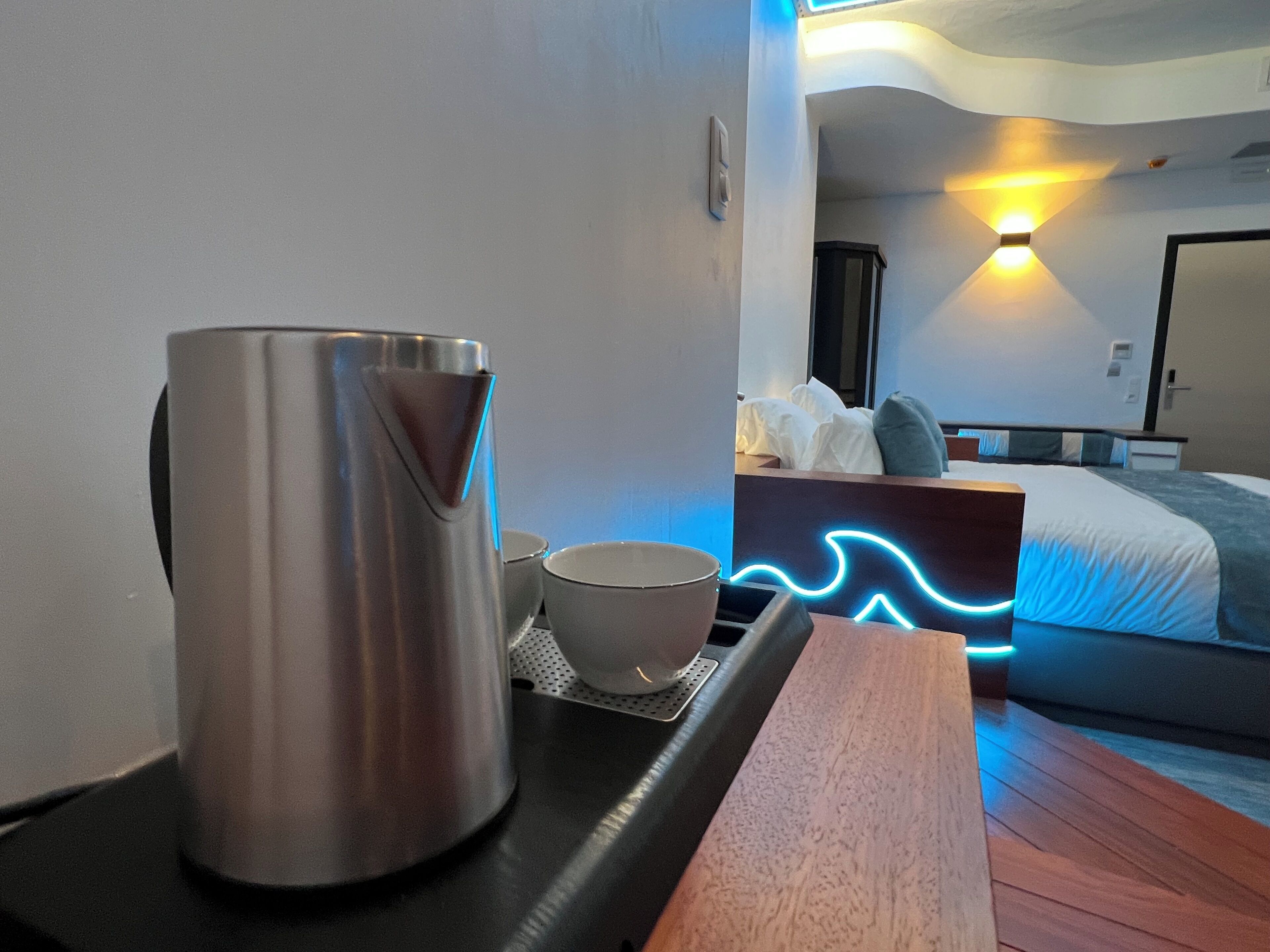 Quarto de hotel moderno com uma chaleira metálica, uma chávena e um tabuleiro sobre uma mesa de madeira. Uma cama com cabeceira iluminada em azul e uma mesa de cabeceira com uma lâmpada são visíveis ao fundo.