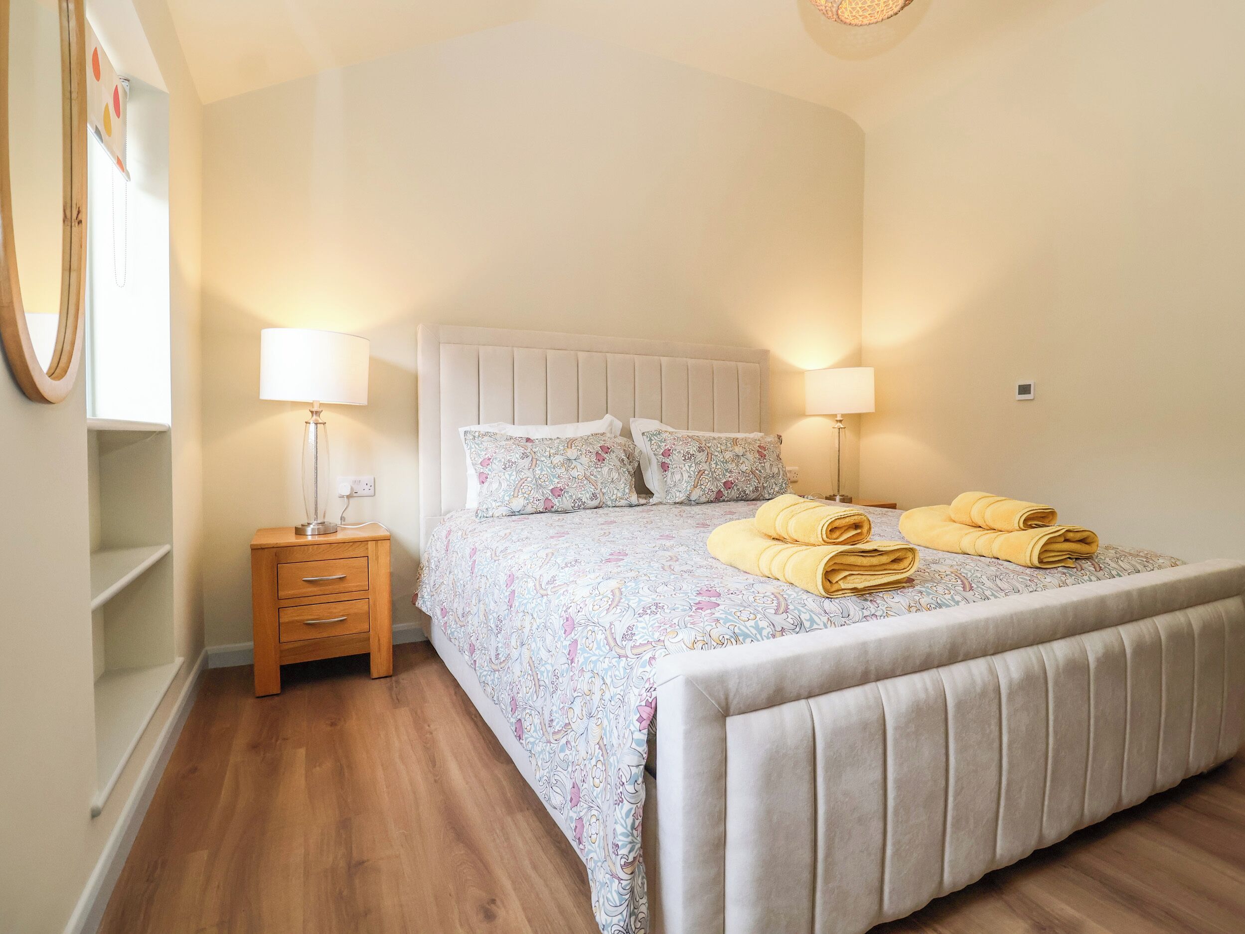 Quarto bem arrumado com cama, abajures, mesa de cabeceira de madeira e um espelho na parede.