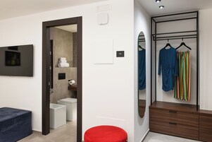 Quarto de hotel moderno com closet espaçoso, um banquinho vermelho e televisão de tela plana.