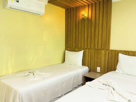 Quarto de hotel com duas camas de solteiro, cabeceira de madeira, ar condicionado fixado na parede e mesa de cabeceira com luminária.