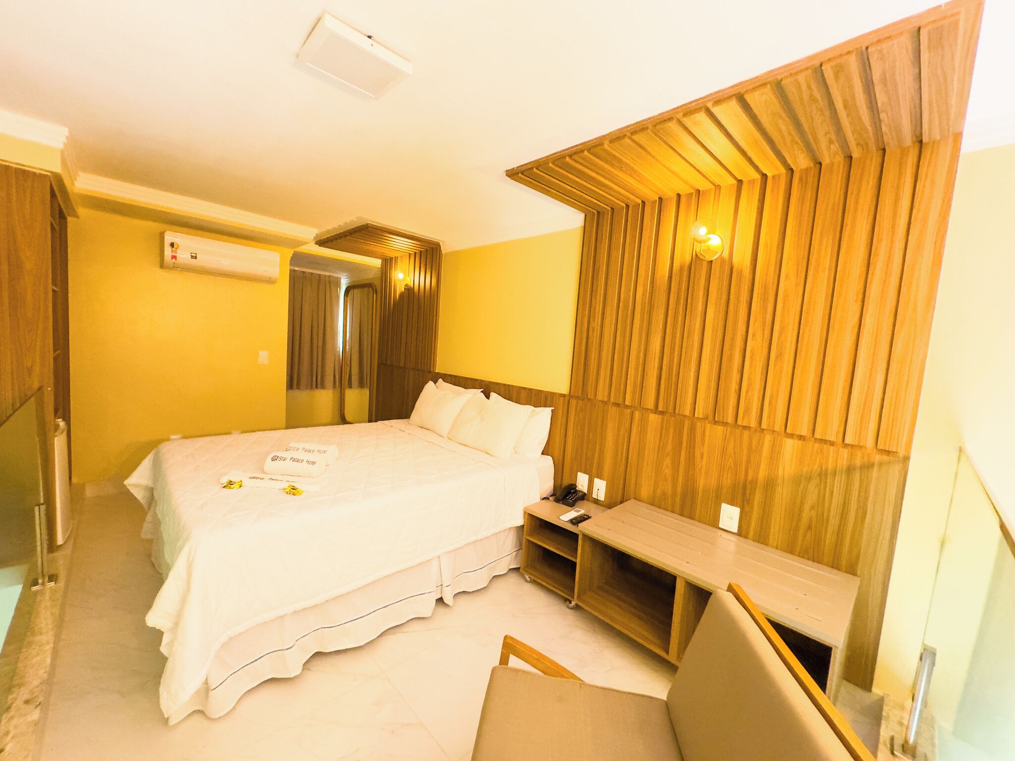 Quarto de hotel com cama, mesa de cabeceira, cadeira e divisória de madeira.