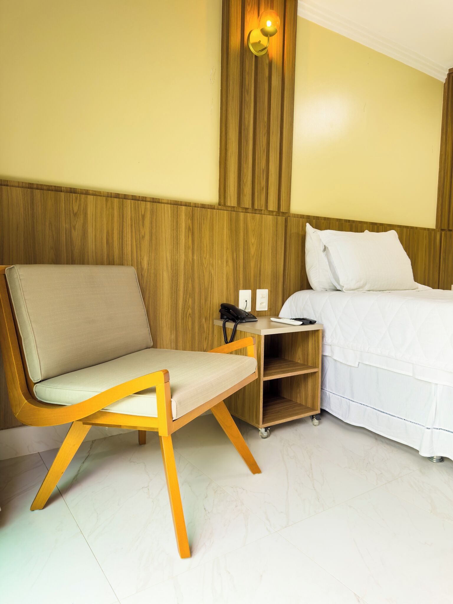 Quarto de hotel com cabeceira de madeira, cama com roupa de cama branca, mesa de cabeceira com telefone e abajur, e uma cadeira com almofada clara.