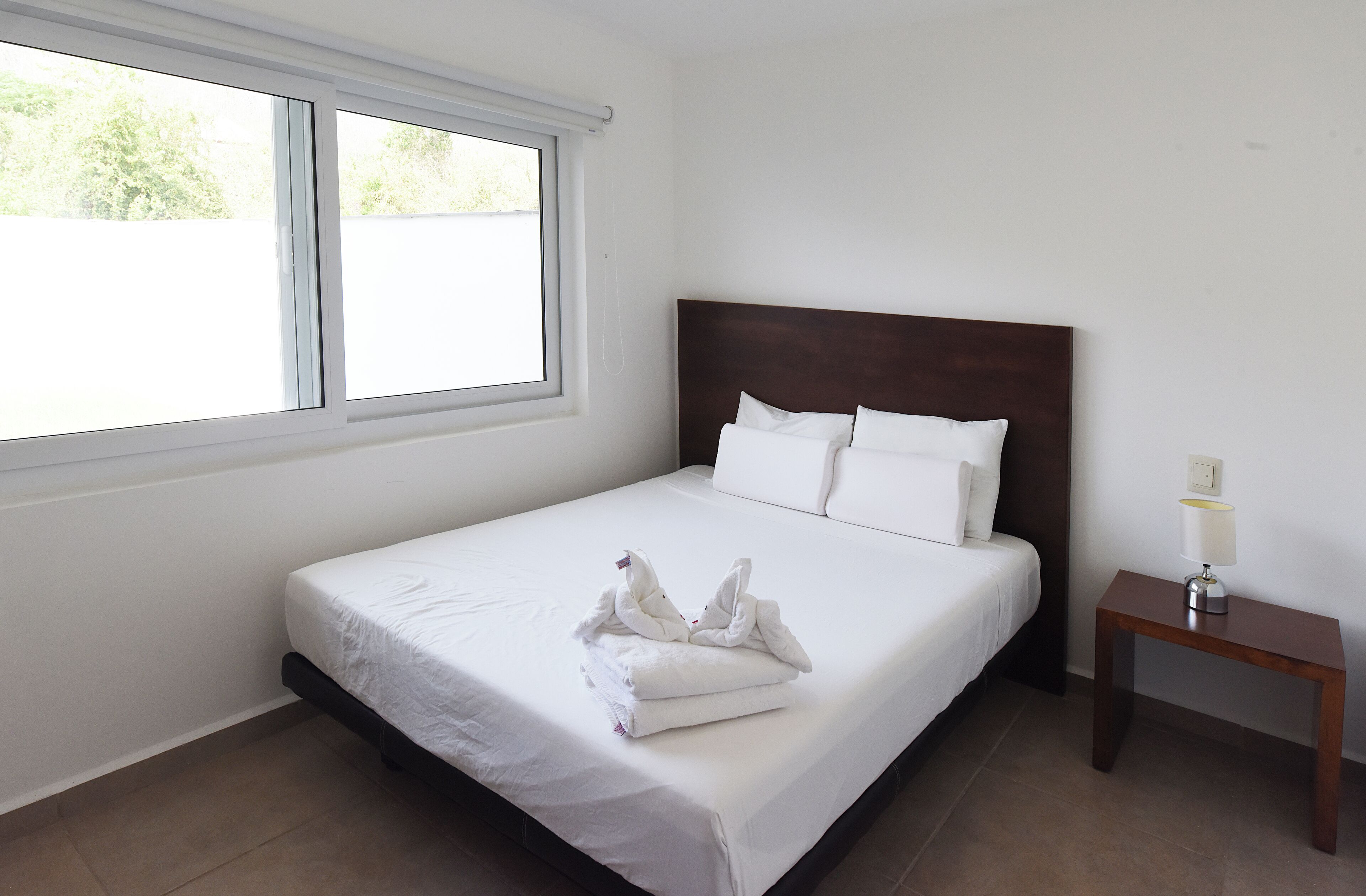 Uma cama bem arrumada com lençóis brancos e cabeceira de madeira, uma mesa de cabeceira com abajur e uma janela com persianas.