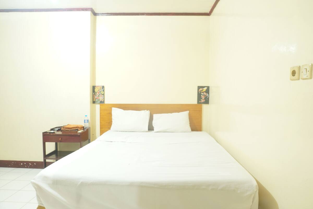 Quarto de hotel com cama, duas almofadas, mesa de cabeceira com garrafa de água e cabeceira de madeira.