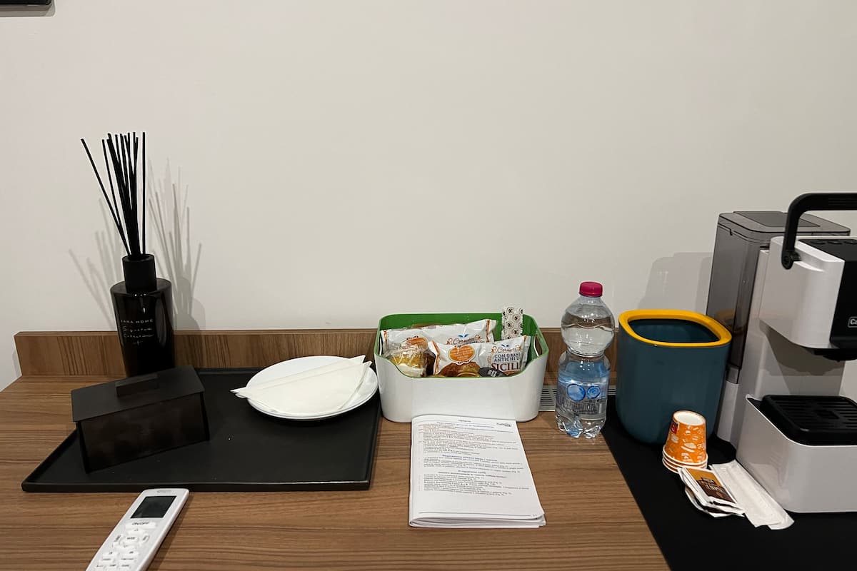 Mesa de quarto de hotel com controle remoto, cardápio, garrafa de água, caixa de lenços, rolo de lenços, xícara, lixeira pequena e uma cesta verde com lanches.