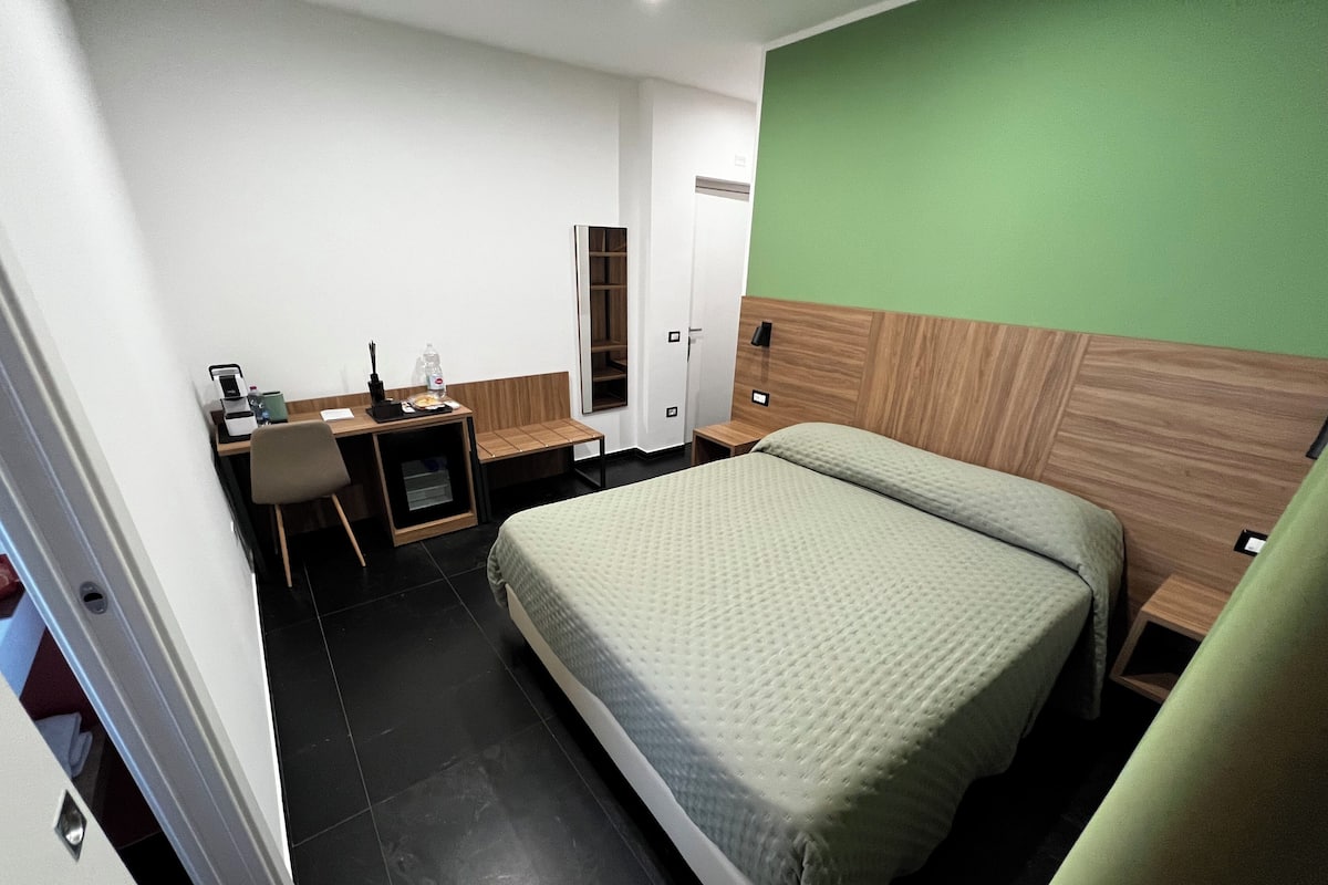Quarto de hotel com cama, escrivaninha com cadeira, mesa pequena, guarda-roupa e uma parede verde decorativa.