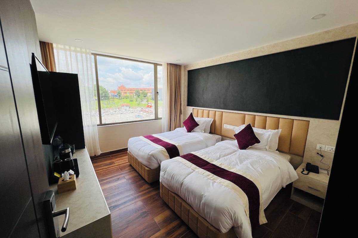 Quarto de hotel com duas camas, televisão e uma janela ampla com vista para edifícios e um estacionamento.