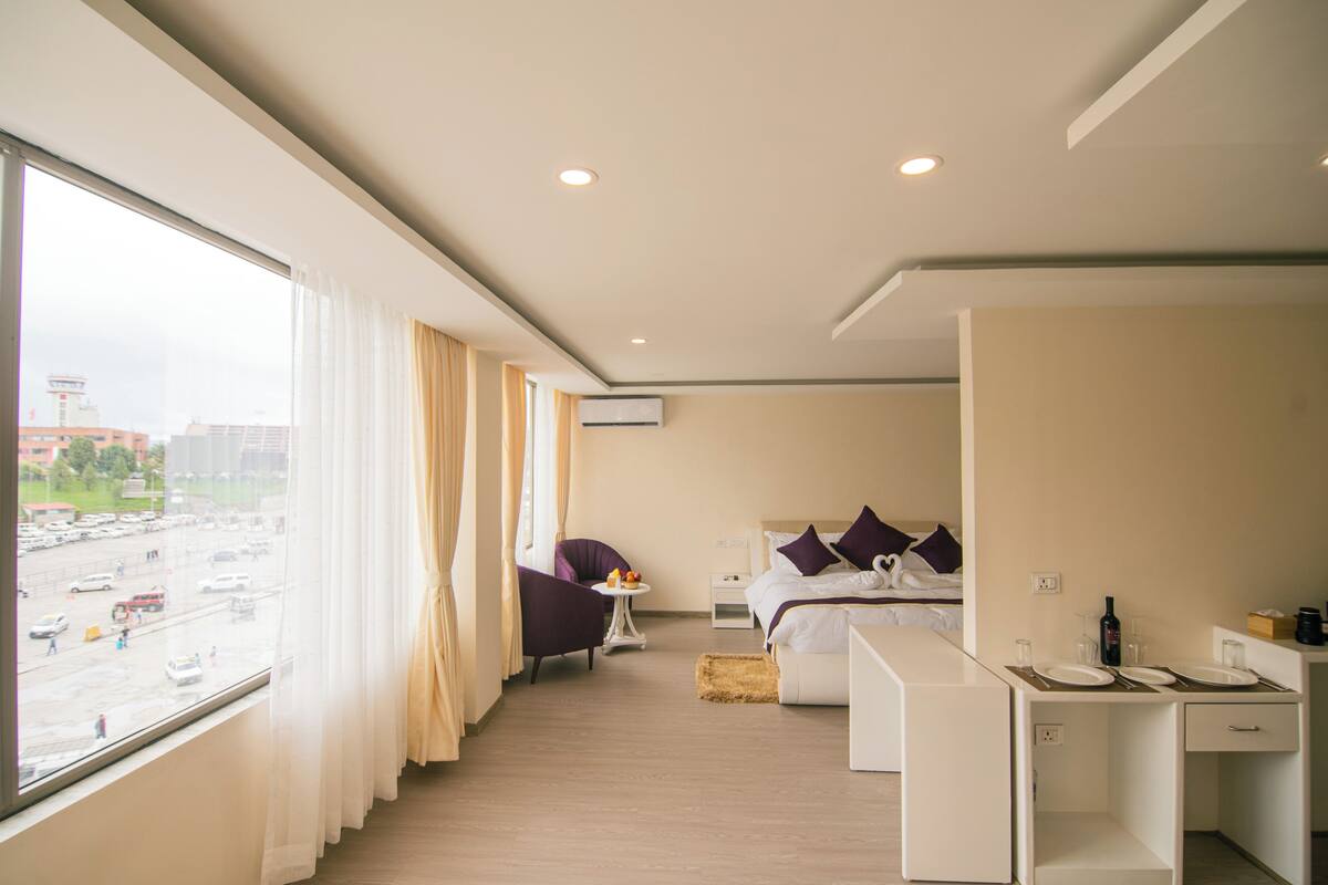 Quarto de hotel com cama, escrivaninha, cadeira, mesinha e vista para uma rua movimentada.