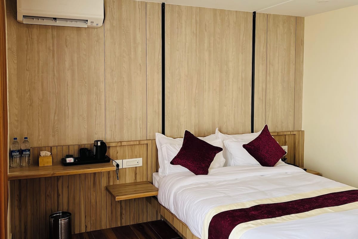 Quarto de hotel com parede de painel de madeira, cama com roupa de cama branca e duas almofadas vermelhas escuras, mesa de cabeceira com cafeteira e garrafas, e um cesto de lixo.