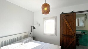 Quarto com cama, luminária de cabeceira, janela e porta de madeira que dá para um banheiro.