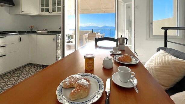 Uma cozinha com armários brancos, uma mesa de jantar com croissants, uma cafeteira e vista para o mar.