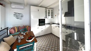 Uma cozinha compacta com armários brancos, um espaço de refeições com mesa de madeira e cadeiras azuis, e um piso estampado.