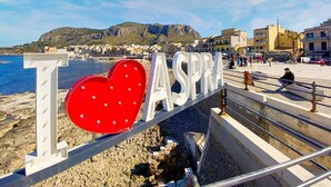 Um letreiro com 'I ♥ ASPRA' e um coração vermelho, com uma vila costeira e montanhas ao fundo.