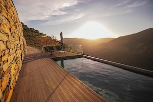 Piscina com deck de madeira e parede de pedra, com vista para uma paisagem montanhosa ao pôr do sol.