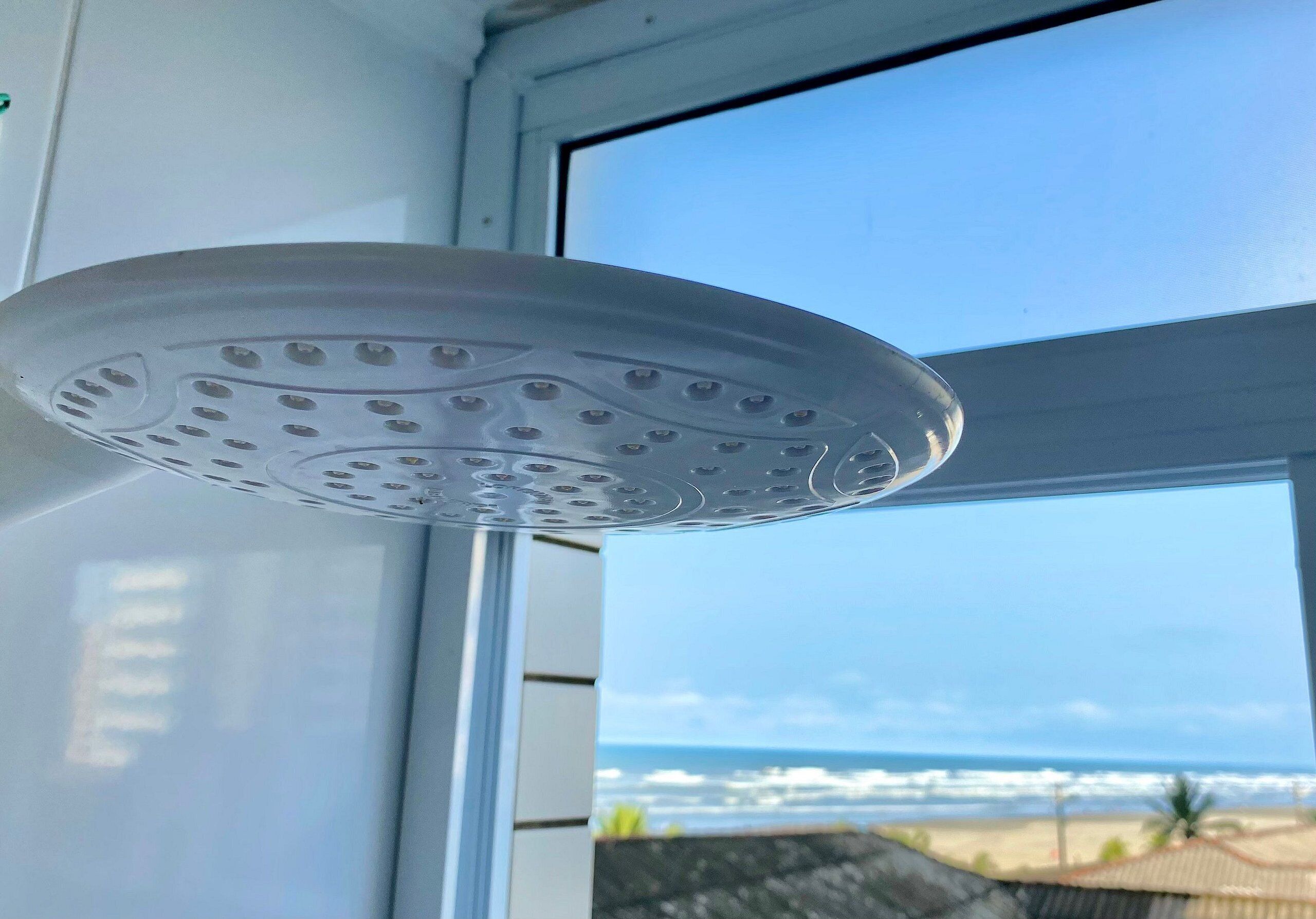 Chuveiro embutido no teto com design circular, janela com vista para a praia e parede revestida de azulejos.