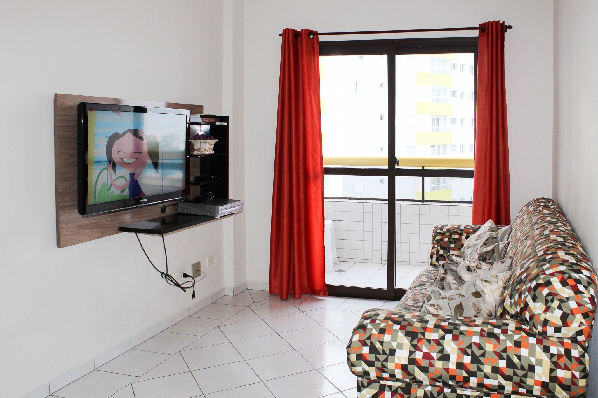 Uma sala de estar com televisão, sofá, janela com cortinas vermelhas e varanda.