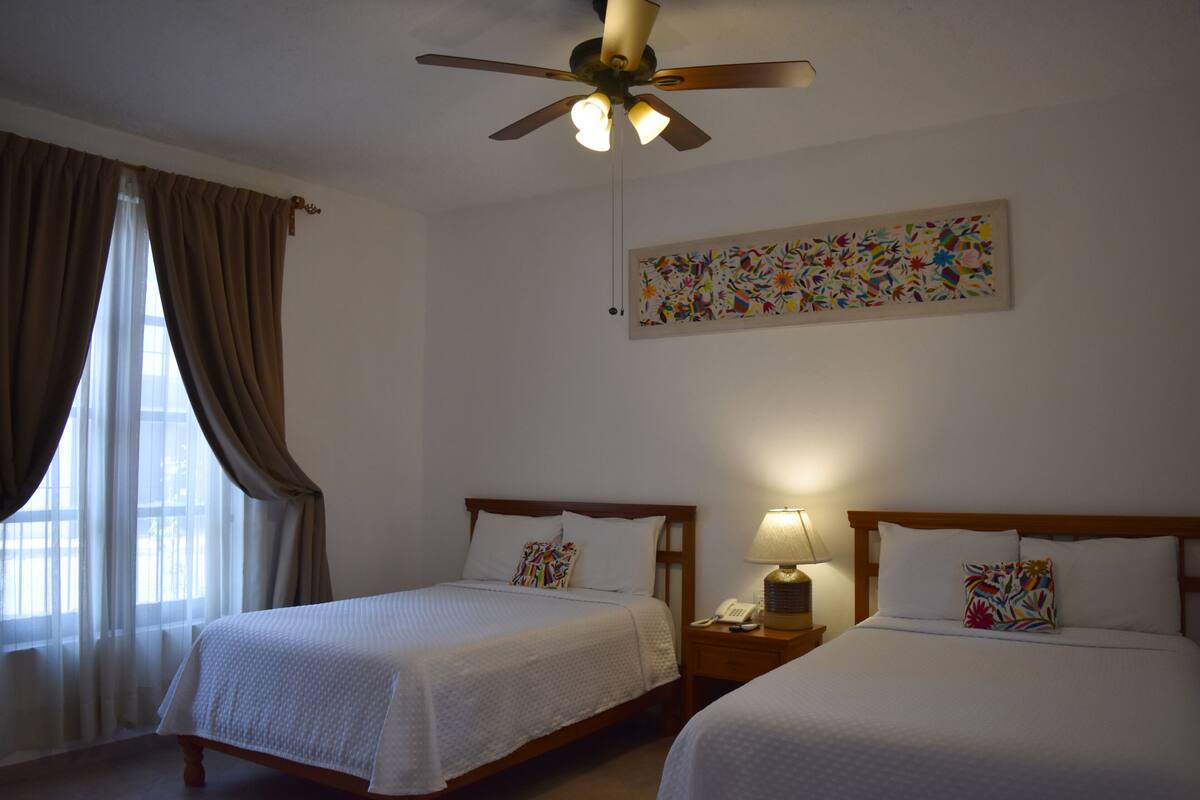 Quarto de hotel com duas camas, um ventilador de teto, um quadro na parede e uma janela com cortinas.