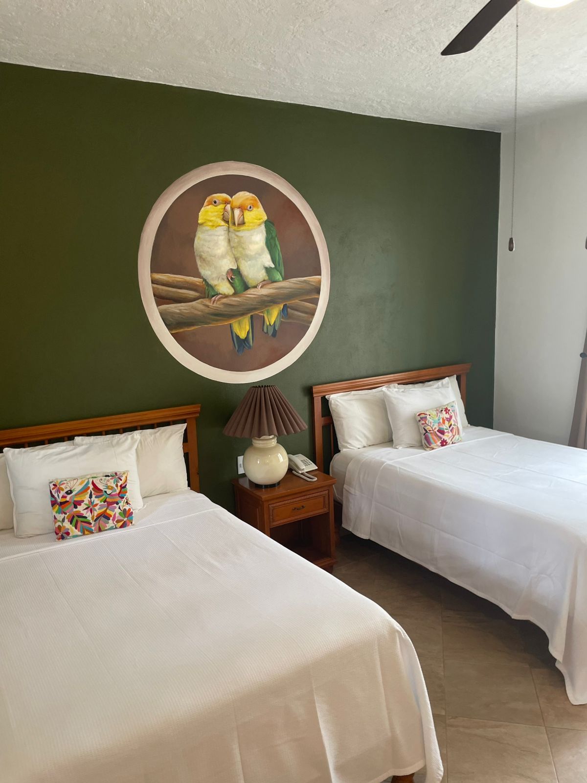 Quarto de hotel com duas camas, uma mesa de cabeceira com abajur e um quadro de pássaros na parede.