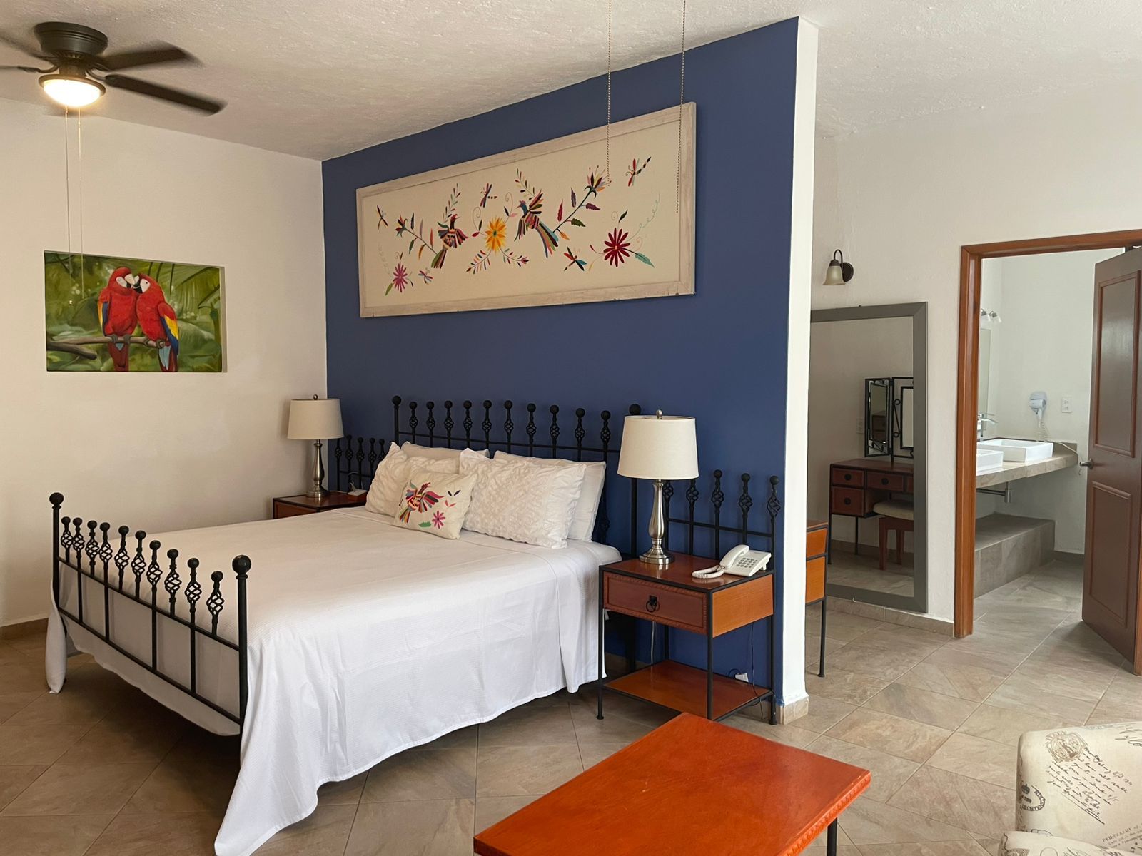 Quarto com cama, mesas de cabeceira, luminária, pintura e banheiro visível por uma porta aberta.