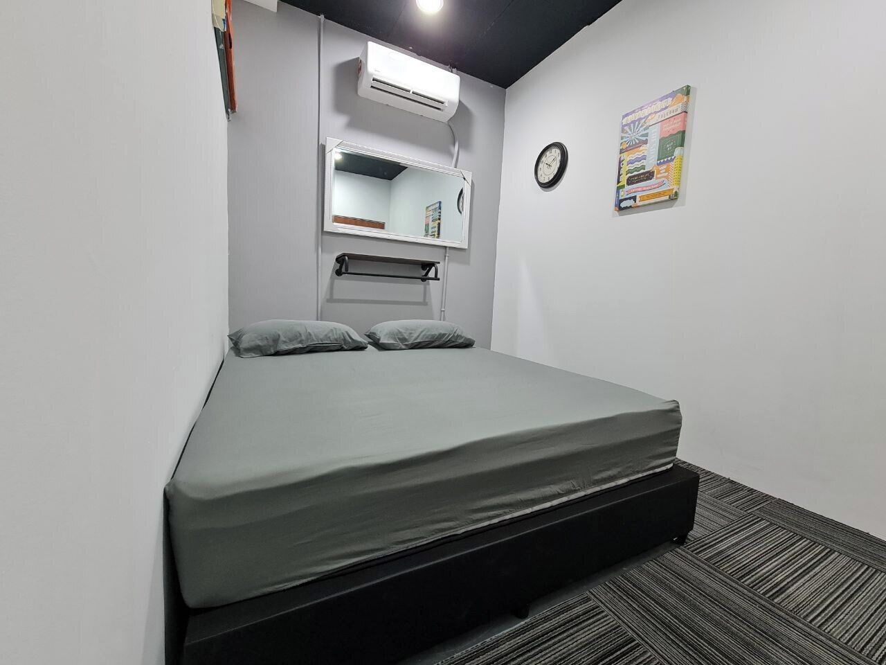 Uma cama de solteiro com colchão e almofadas, ar condicionado fixado na parede, uma pequena prateleira com espelho e um relógio na parede.