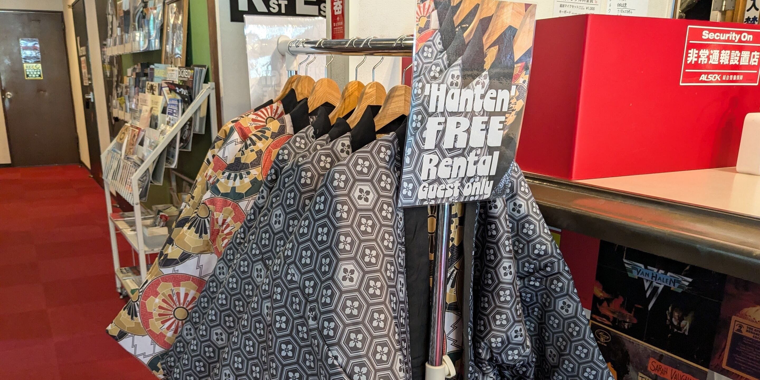 Um cabideiro com diversas peças de roupa estampadas, uma placa indicando 'Hanten Free Rental' e uma placa de segurança vermelha.
