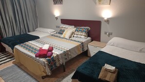 Quarto de hotel com duas camas, uma cama grande com colcha estampada e almofadas, e uma cama menor com colcha lisa. Há duas luminárias fixadas na parede, uma janela com cortinas e uma pequena mesa com uma toalha.