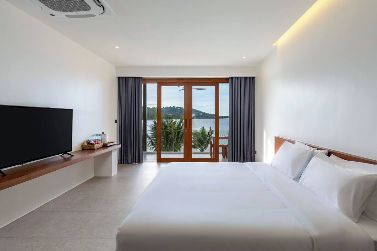 Quarto de hotel com uma cama grande, uma televisão de tela plana em um console de madeira, uma varanda com vista para o mar e uma porta de vidro deslizante.