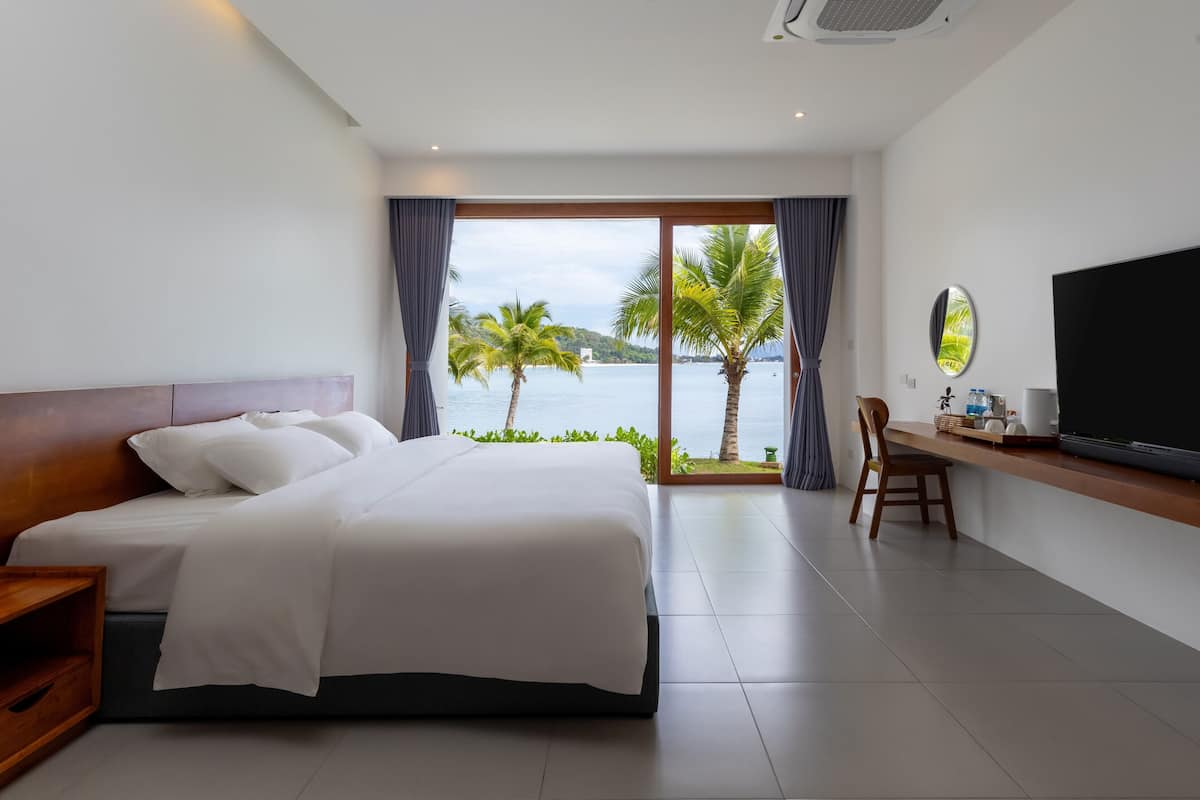 Quarto de hotel com uma cama grande, uma mesa de trabalho com televisão e vista para o mar e palmeiras.