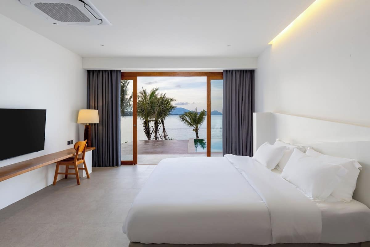 Quarto de hotel com uma cama grande, uma mesa e uma televisão. Vê-se o mar e palmeiras pela janela.