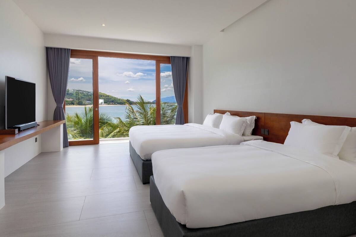 Quarto de hotel com duas camas, televisão e uma grande janela com vista para o mar.