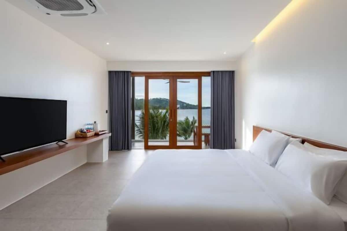 Quarto de hotel com uma cama grande, uma TV sobre uma prateleira de madeira, e vista para o mar através de uma porta de vidro.