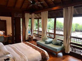 Quarto com cama, área de estar com almofadas, escrivaninha e janelas amplas com cortinas.