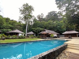 Uma área de piscina com espreguiçadeiras e guarda-sóis, cercada por vegetação exuberante e cabanas de estilo tradicional.