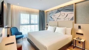 Quarto de hotel com uma cama grande, uma mesa com um controle remoto, uma cadeira, mesa de cabeceira com telefone e janela com cortinas.