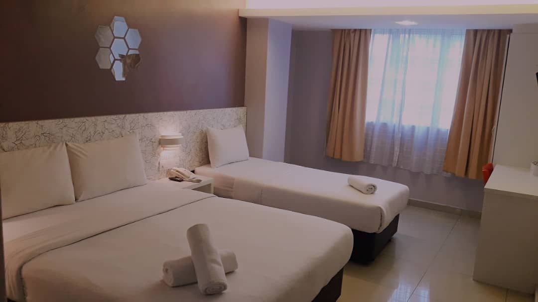 Quarto de hotel com duas camas, mesa de cabeceira com telefone, luminária na parede e janela com cortinas.